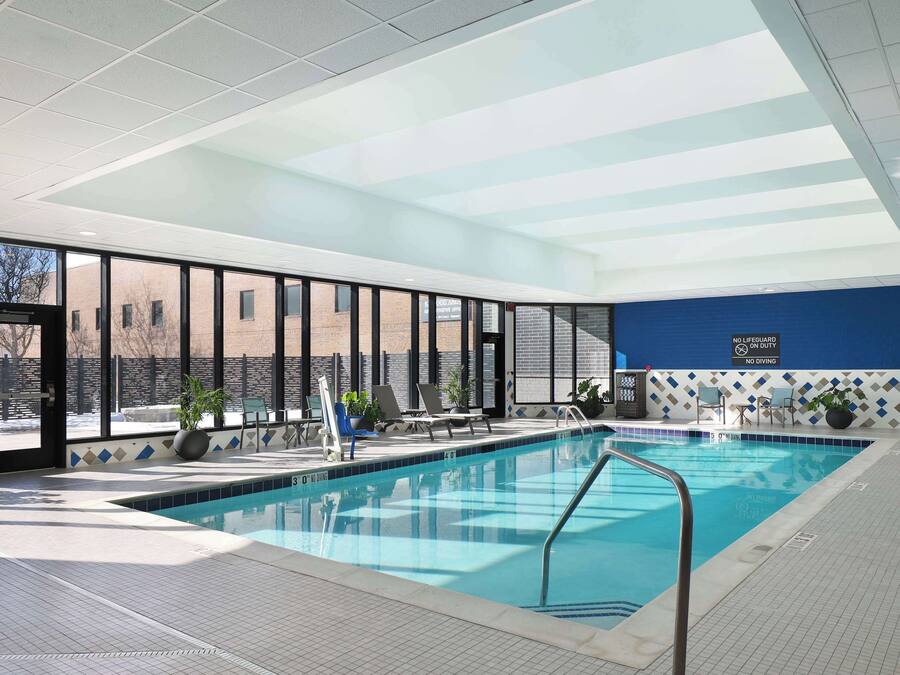 Piscina interna com paredes de vidro transparente, piso revestido e parede azul com aviso 'No Lifeguard, No Diving'.