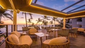 Um restaurante à beira-mar com mobiliário de vime, mesas e vista para o oceano ao pôr do sol.