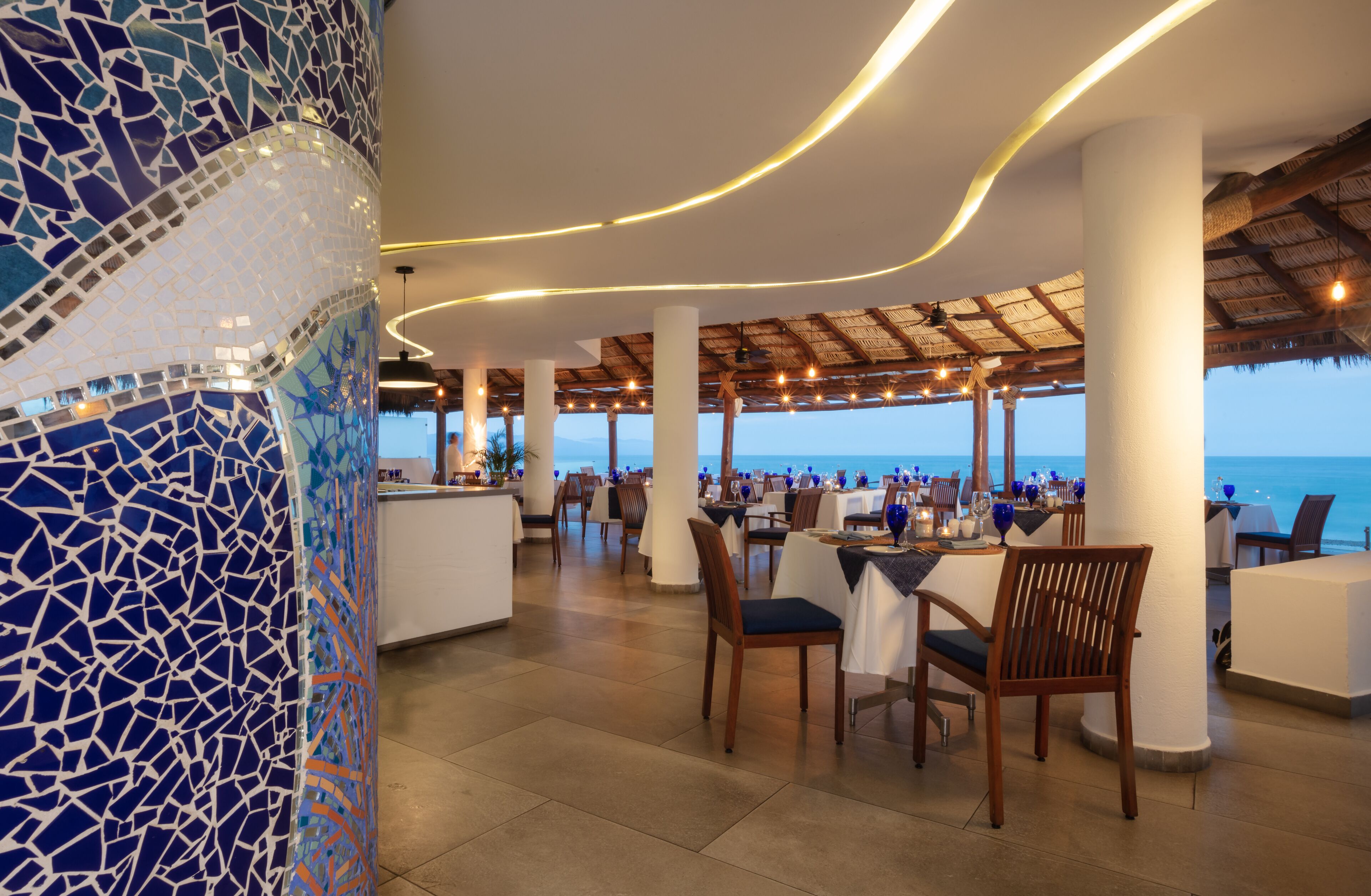 Um espaço de refeições interno com mesas e cadeiras, uma parede decorativa com mosaicos e vista para o mar.