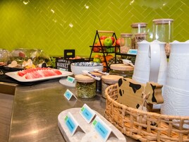 Uma área de buffet com diversos alimentos, incluindo frutas e acompanhamentos, e recipientes de servir identificados.
