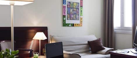 Quarto de hotel com cama, escrivaninha com laptop, luminária, planta e uma obra de arte na parede.