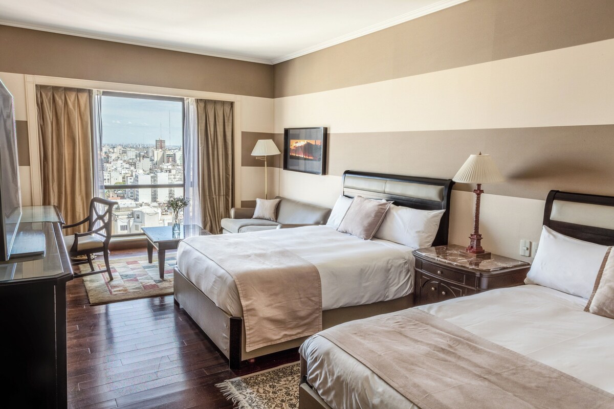 Quarto de hotel com duas camas, uma escrivaninha, uma cadeira, janela com cortinas, vista para a cidade e mesa de cabeceira com abajur.