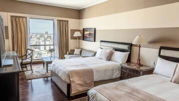 Quarto de hotel com duas camas, uma escrivaninha, uma cadeira, janela com cortinas, vista para a cidade e mesa de cabeceira com abajur.