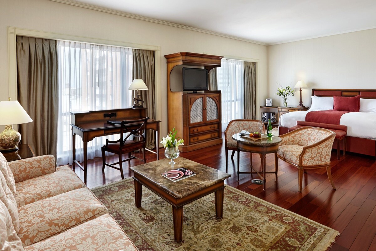 Quarto de hotel com sofá, mesa de centro, escrivaninha com piano, televisão, cama e área de refeições com cadeiras.