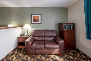 Quarto com sofá de couro marrom, escrivaninha pequena com luminária, quadro pendurado na parede e micro-ondas em armário.