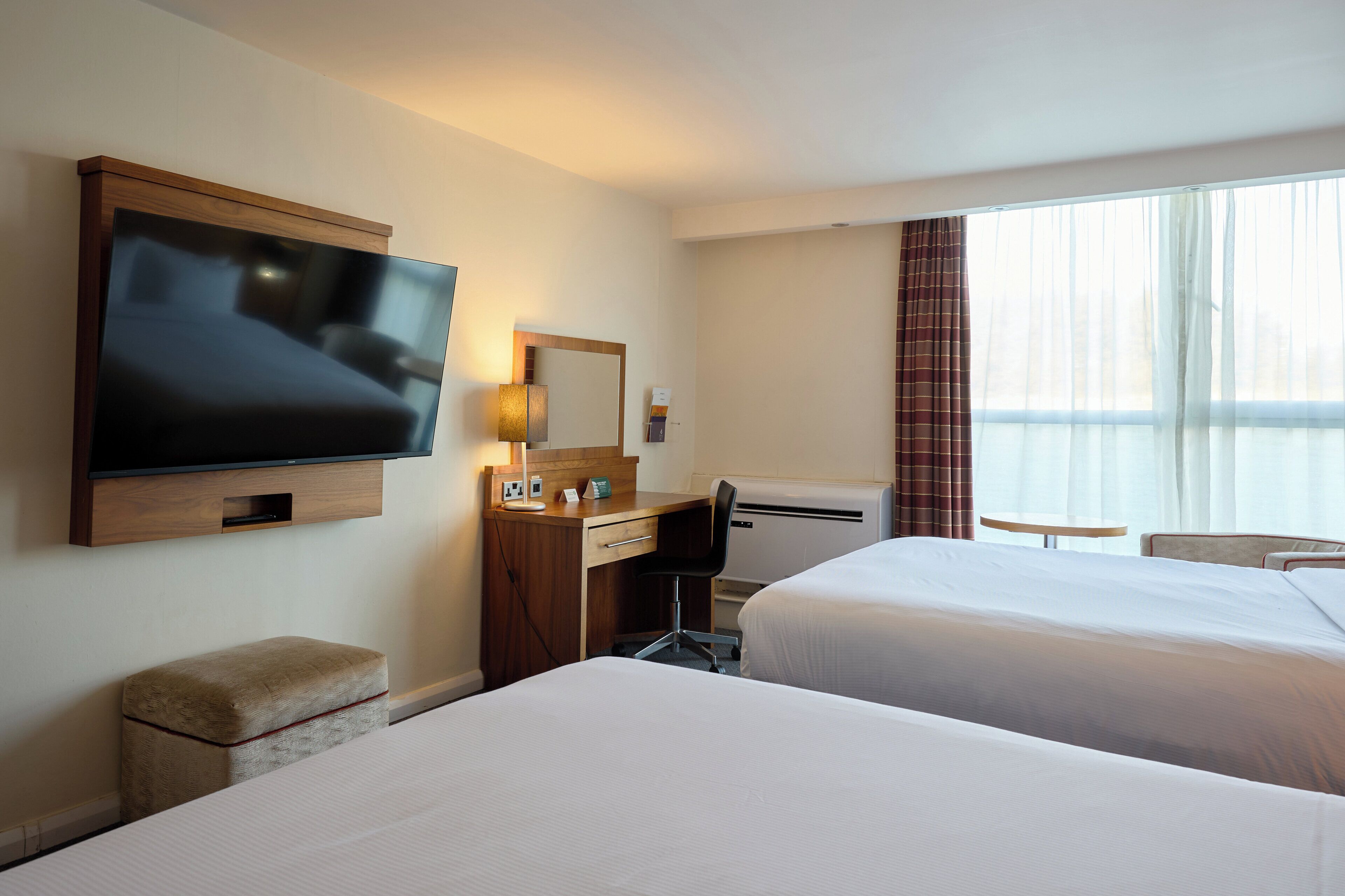 Quarto de hotel com duas camas, uma escrivaninha, uma cadeira, uma televisão e uma janela com cortinas.