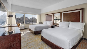 Quarto de hotel com duas camas, uma escrivaninha, uma cadeira e uma janela grande com vista para a cidade.