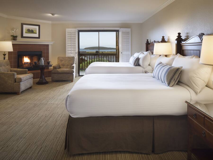 Quarto de hotel com duas camas, lareira, varanda com vista e área de estar com cadeiras.