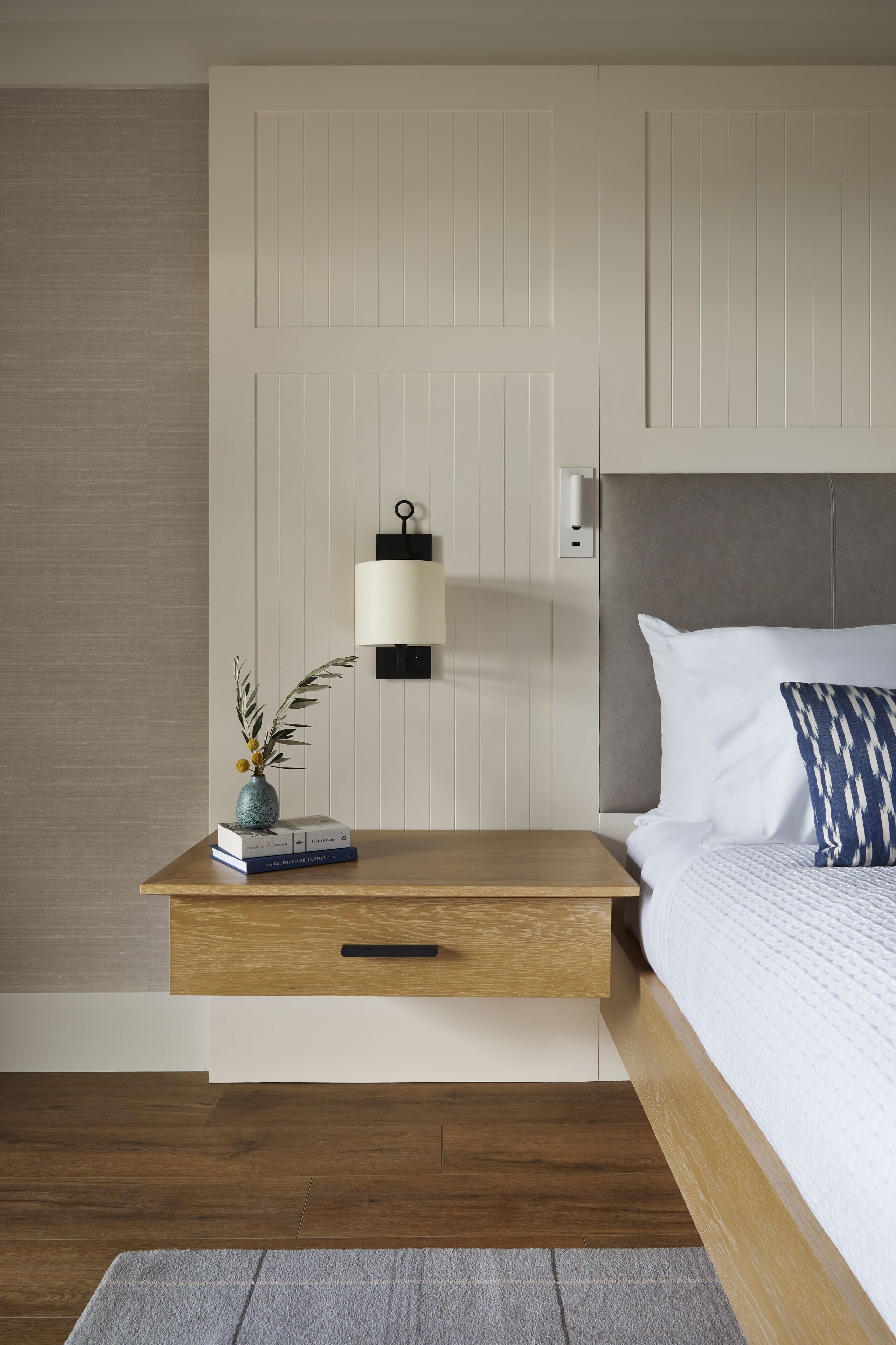 Quarto com mesa de cabeceira de madeira, luminária, cama com roupa de cama branca e tapete cinza.