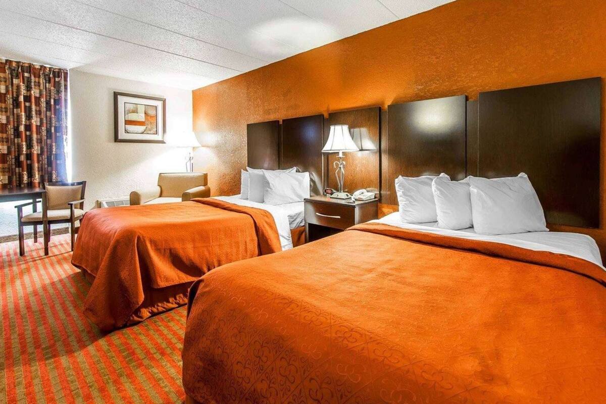 Quarto de hotel com duas camas, uma escrivaninha, uma cadeira e um abajur.