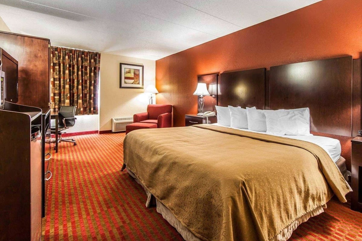 Quarto de hotel com uma cama grande, uma escrivaninha com cadeira, uma mesinha de apoio com luminária e uma poltrona vermelha.