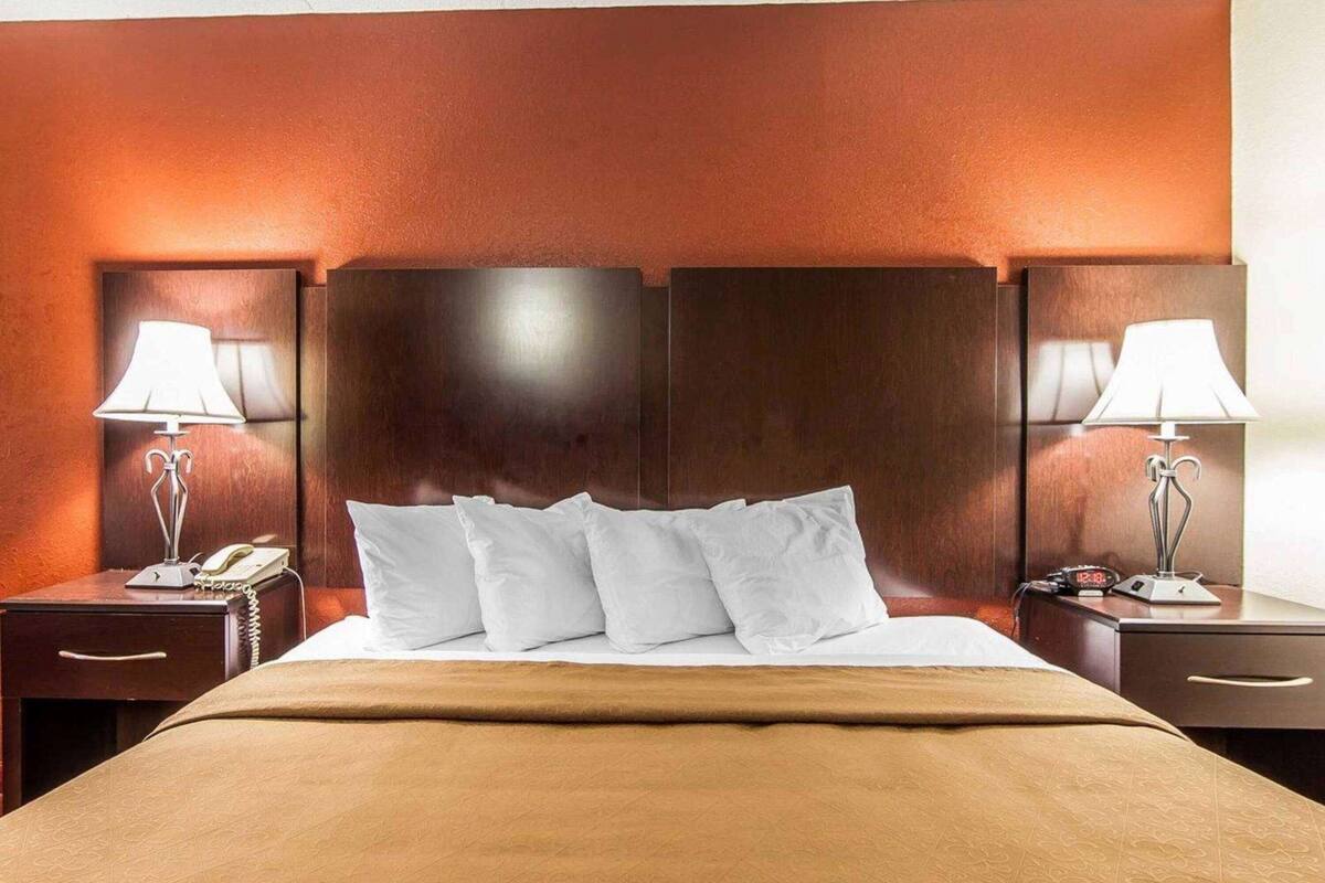 Quarto de hotel com uma cama grande, duas mesas de cabeceira com abajures, um telefone e um relógio em uma das mesas.