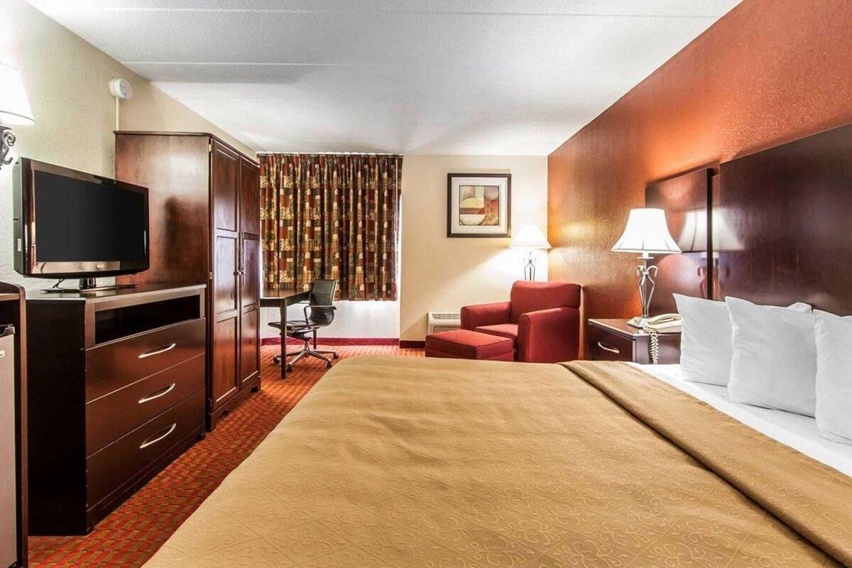 Quarto de hotel com cama, escrivaninha com televisão, cadeira, luminária de mesa e um quadro na parede.