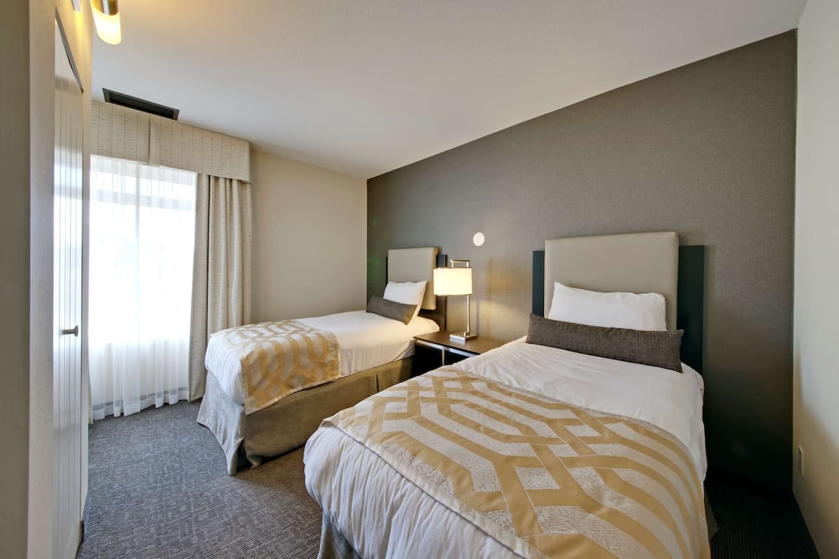 Quarto de hotel com duas camas, janela com cortinas, mesa de cabeceira com abajur e luminária na parede.
