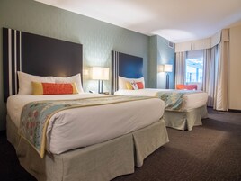 Quarto de hotel com duas camas, cada uma com cabeceira, mesas de cabeceira, luminárias e uma janela com cortinas.