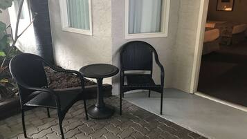 Uma área de estar nos cantos, com duas cadeiras, uma mesa pequena, uma janela e uma porta que dá para um quarto.