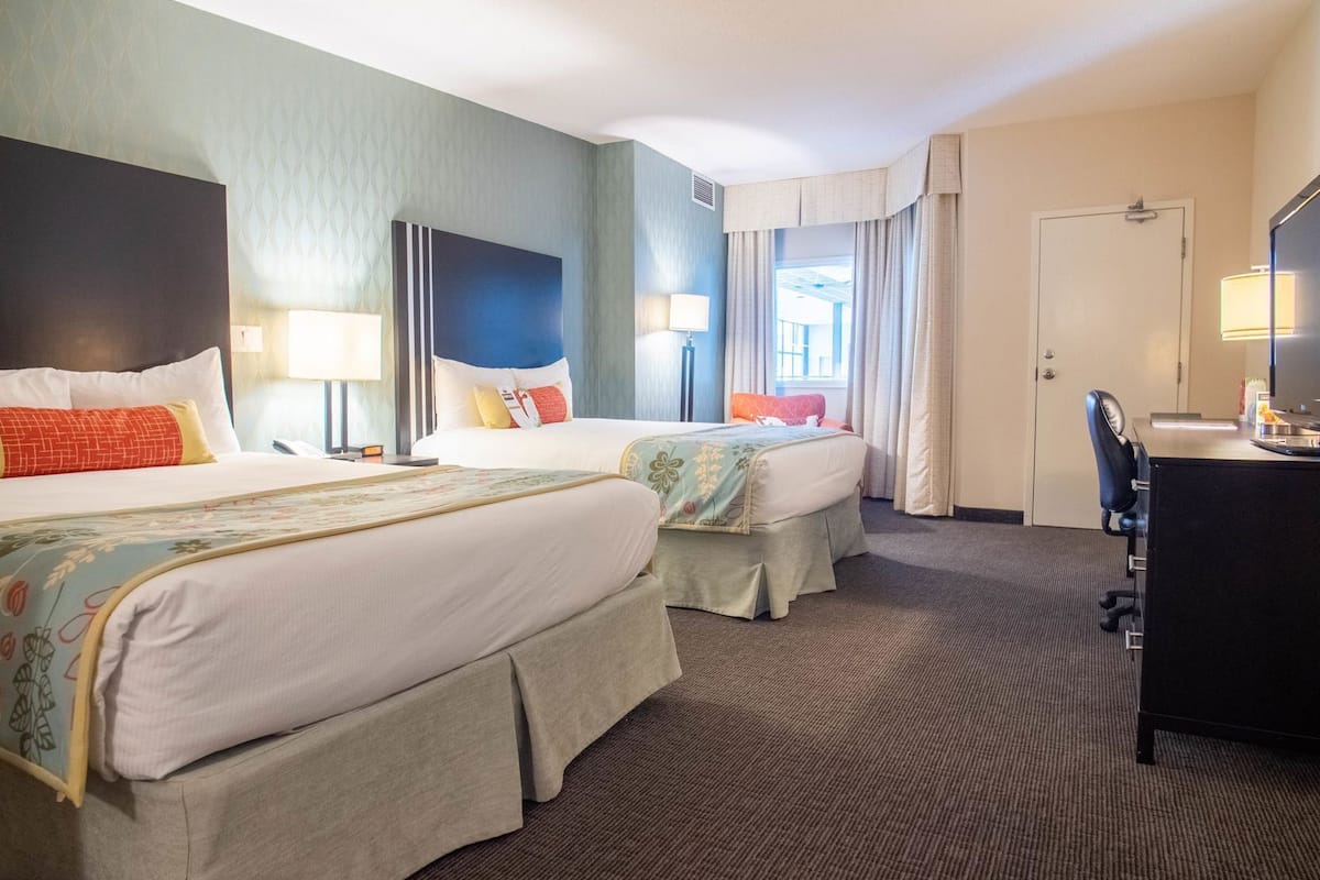 Quarto de hotel com duas camas, uma escrivaninha, uma cadeira, uma televisão e uma porta.
