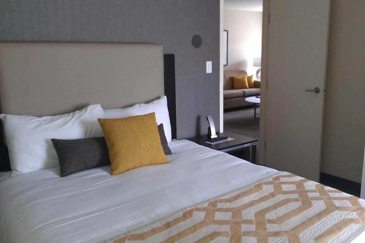 Quarto de hotel com cama, mesa de cabeceira, luminária e porta que dá para outro cômodo.