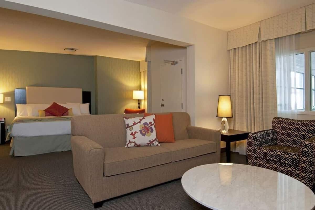 Quarto de hotel com cama, sofá, poltrona, mesa redonda e janela com cortinas.