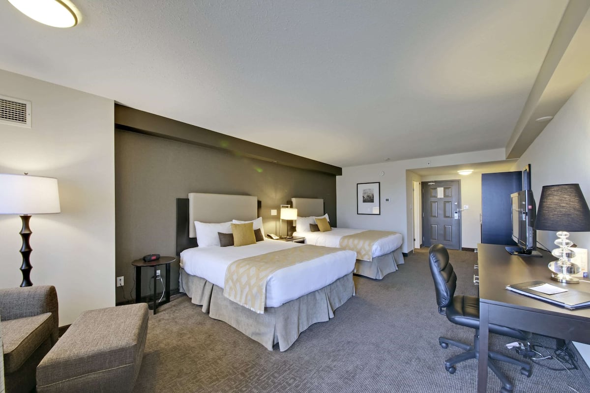 Quarto de hotel com duas camas, uma escrivaninha, uma cadeira, um abajur e uma pequena mesa.