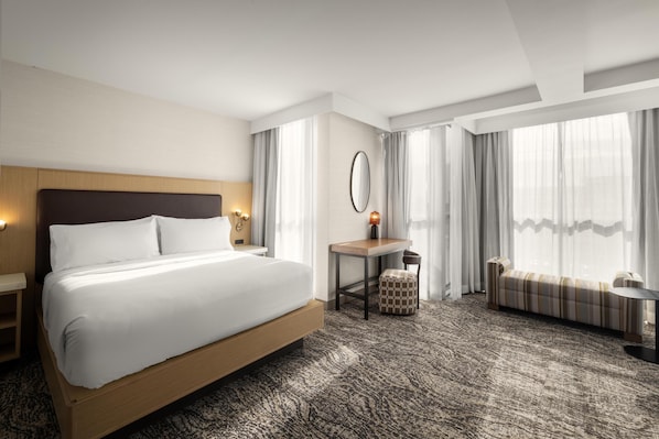 Quarto de hotel com uma cama grande, uma mesa pequena, um sofá e uma janela ampla com cortinas.