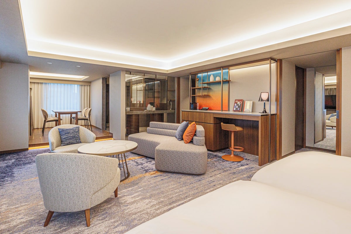 Quarto de hotel moderno com sofá, poltrona e mesa pequena. Há uma escrivaninha com abajur e espelho. O quarto possui uma janela grande com persianas.
