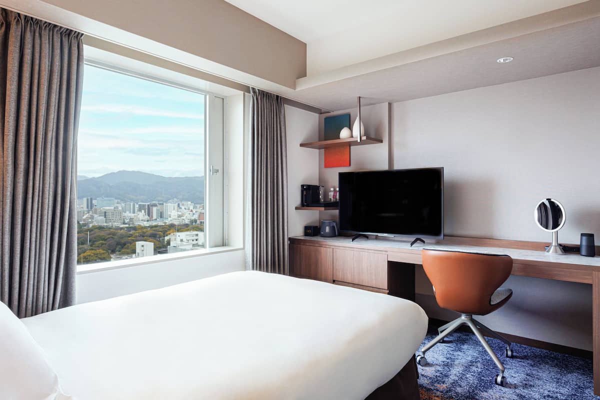 Quarto de hotel com uma cama, uma escrivaninha com computador, televisão e vista para a paisagem urbana através de uma janela grande.