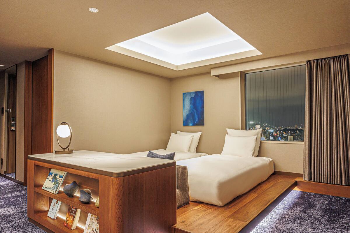 Quarto de hotel com claraboia, cama, mesa de cabeceira com abajur, estante de livros e vista para a cidade pela janela.