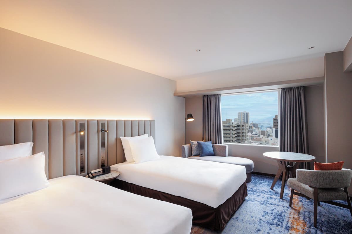 Quarto de hotel com duas camas, uma mesa pequena, uma cadeira e vista para a cidade.