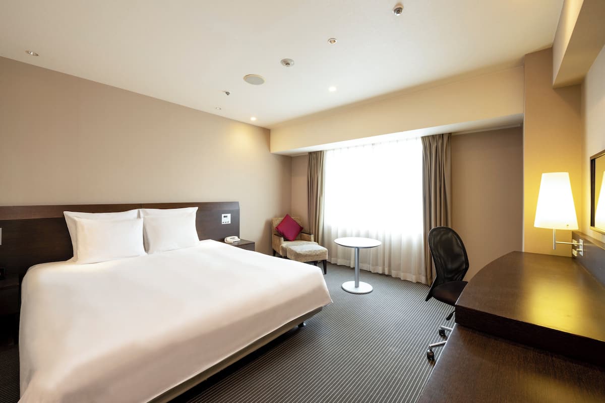 Quarto de hotel com uma cama grande, uma escrivaninha, uma cadeira, uma mesa redonda pequena e uma janela com cortinas.