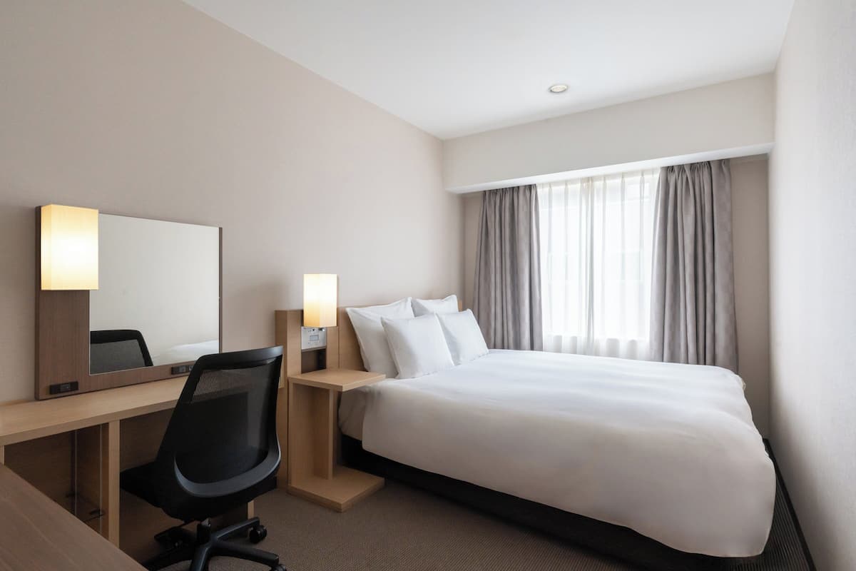 Quarto de hotel com cama, escrivaninha, cadeira, mesa de cabeceira e luminária, além de uma janela grande com cortinas.