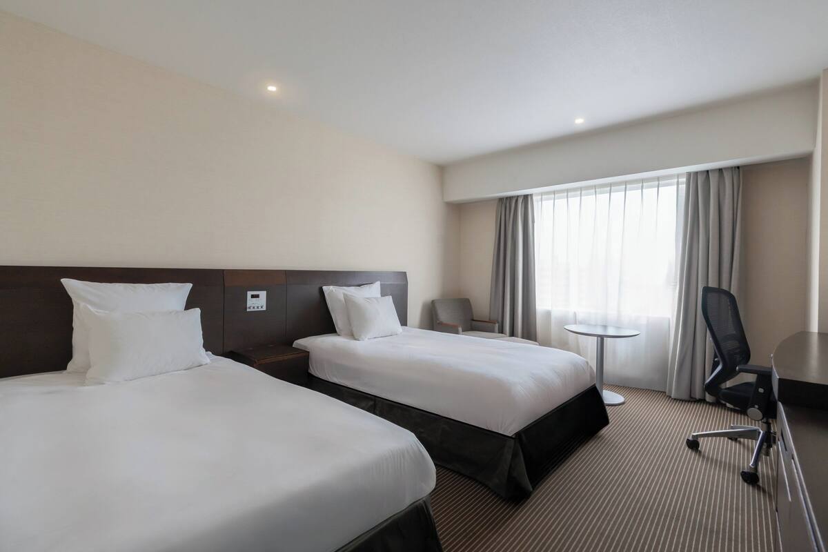 Quarto de hotel com duas camas, uma mesa redonda pequena, uma cadeira e uma janela grande com cortinas.