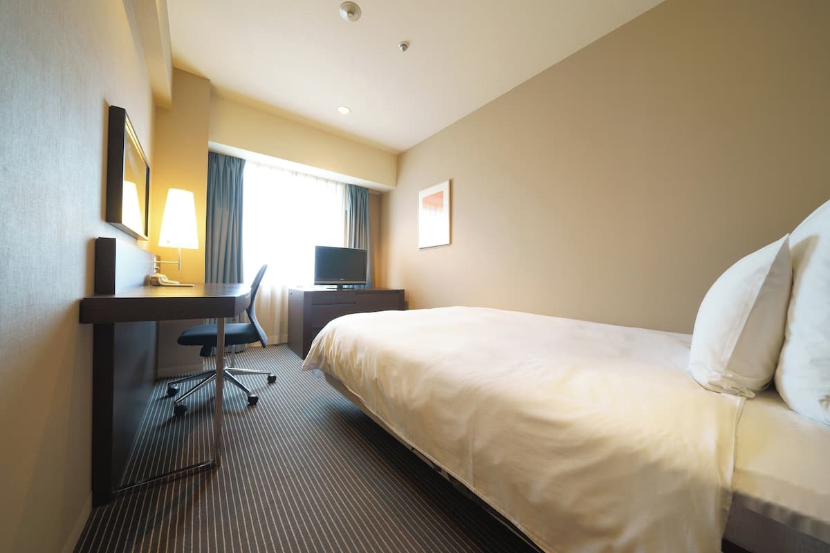Quarto de hotel com cama, escrivaninha, cadeira, abajur e janela com cortinas.
