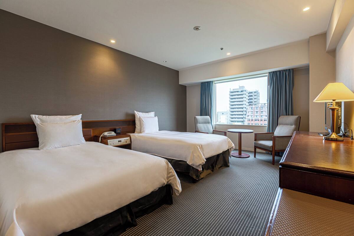 Quarto de hotel com duas camas, uma mesa, uma cadeira, uma luminária e vista para a cidade.