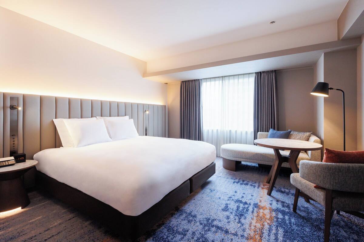 Quarto de hotel moderno com uma cama grande, uma mesa de centro redonda, um sofá e uma escrivaninha com abajur.