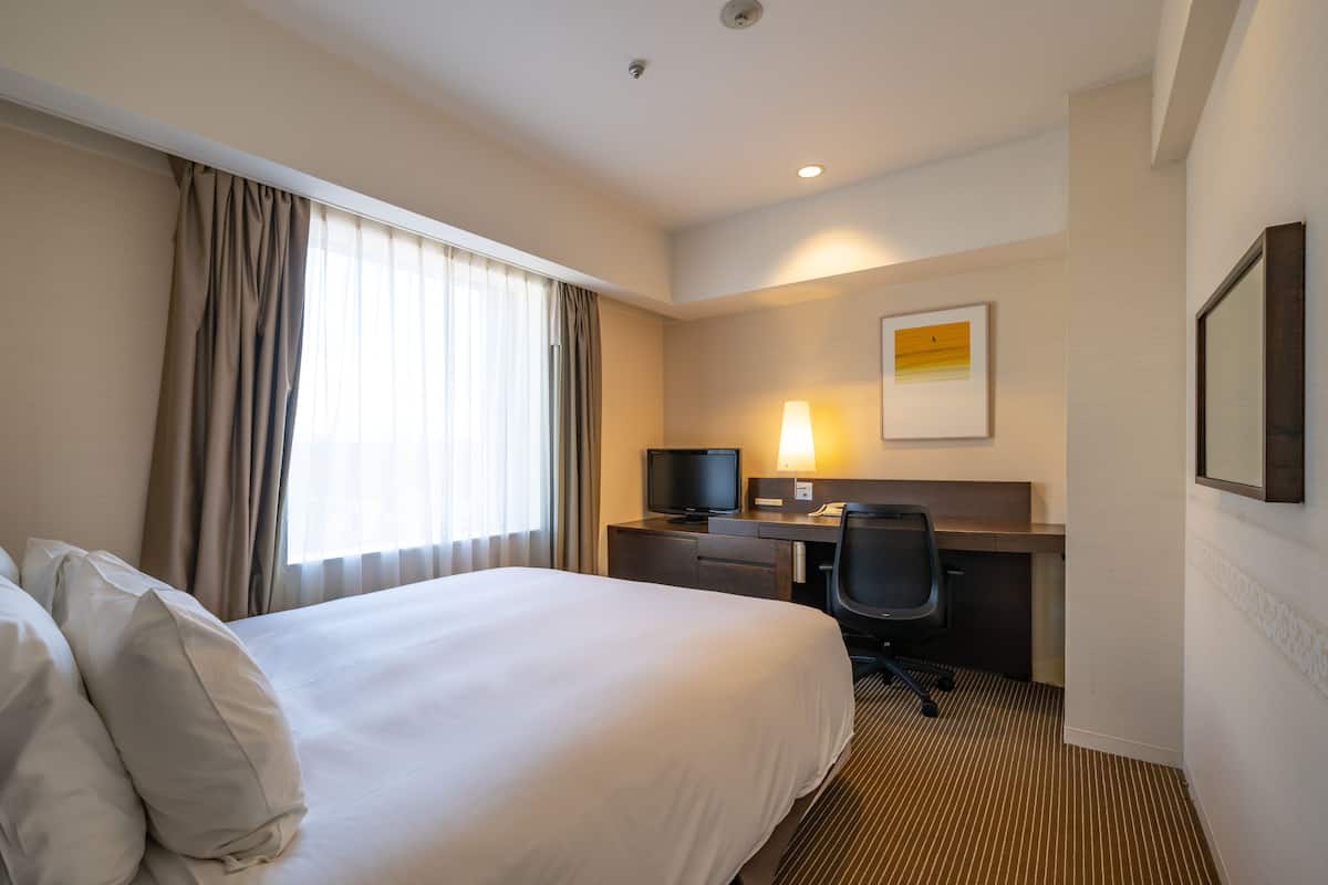 Quarto de hotel com uma cama grande, uma escrivaninha com computador, uma cadeira e uma televisão.