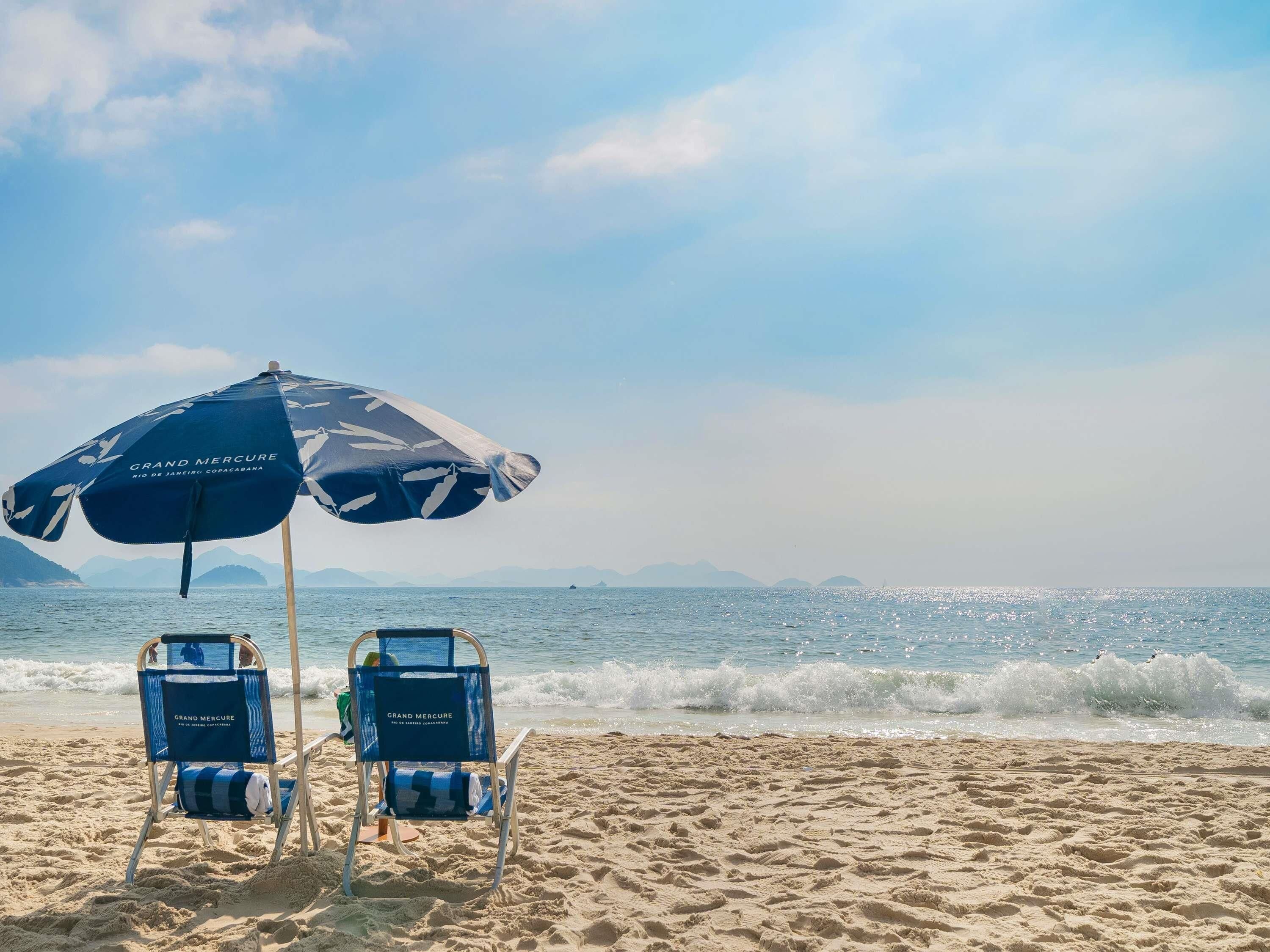 Duas cadeiras de praia com um guarda-sol identificado como 'Grand Mercure Rio de Janeiro Copacabana' em uma praia arenosa, com o oceano e montanhas ao fundo.