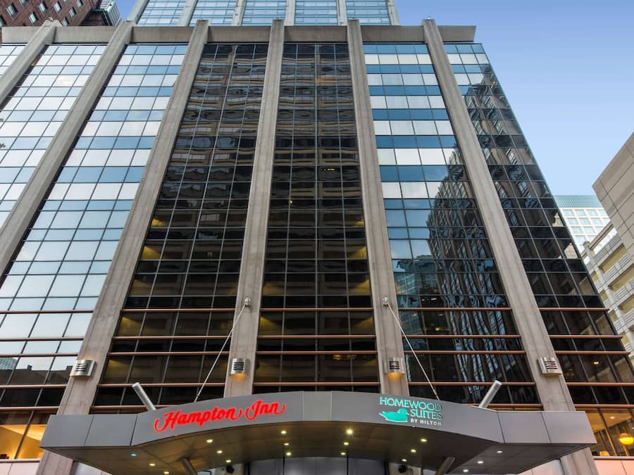 Um hotel com fachada moderna em vidro, com destaque para a placa Hampton Inn e a placa Homewood Suites by Hilton.