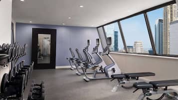 Academia com equipamentos de cardio, halteres e um banco. Possui uma grande janela com vista para a cidade.