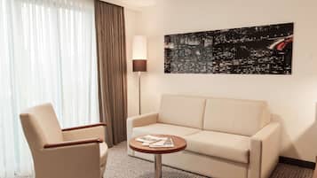 Quarto de hotel com sofá bege, mesa lateral de madeira com revistas, luminária de chão e pintura de paisagem urbana na parede.