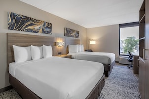 Quarto de hotel com duas camas, uma escrivaninha, uma cadeira e uma janela grande.