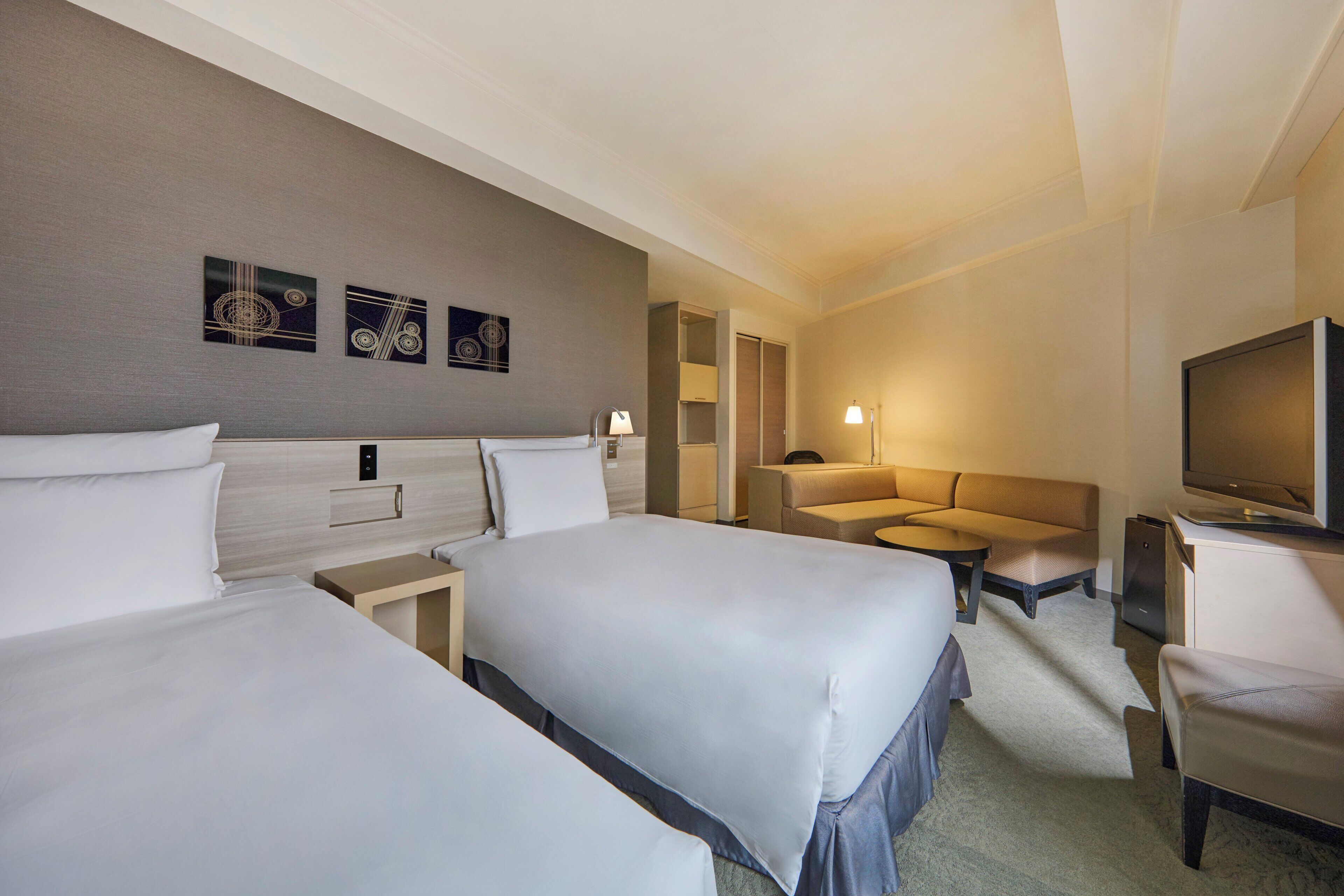 Quarto de hotel com duas camas, um sofá, uma televisão e uma escrivaninha.
