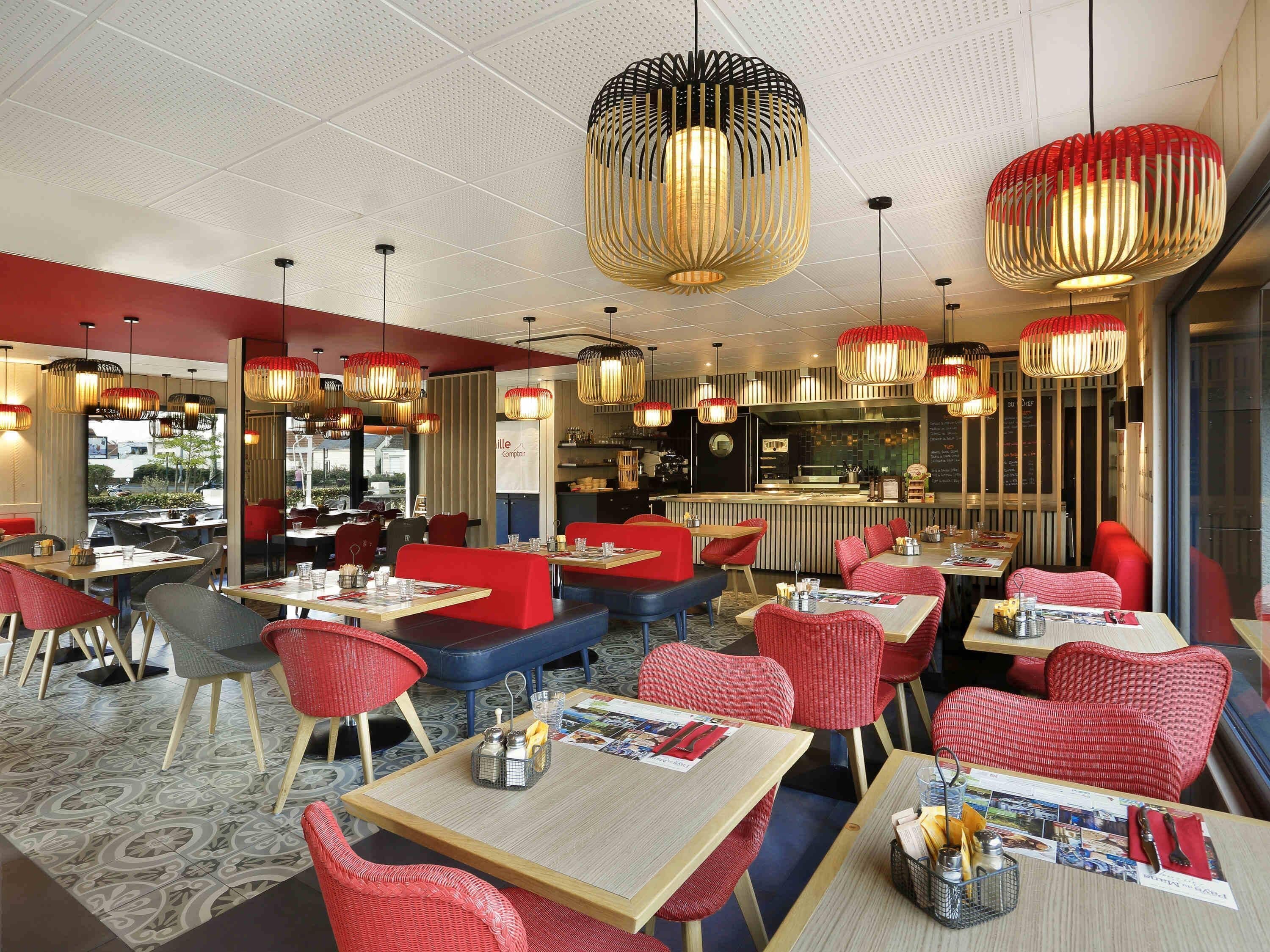 Interior de restaurante com assentos vermelhos e azuis, várias luminárias pendentes e uma área de bar visível através do separador de vidro.