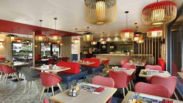 Interior de restaurante com assentos vermelhos e azuis, várias luminárias pendentes e uma área de bar visível através do separador de vidro.