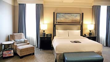 Quarto de hotel com uma cama grande, uma cadeira, uma mesa pequena e um banquinho.