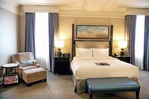 Quarto de hotel com uma cama grande, uma cadeira, uma mesa pequena e um banquinho.