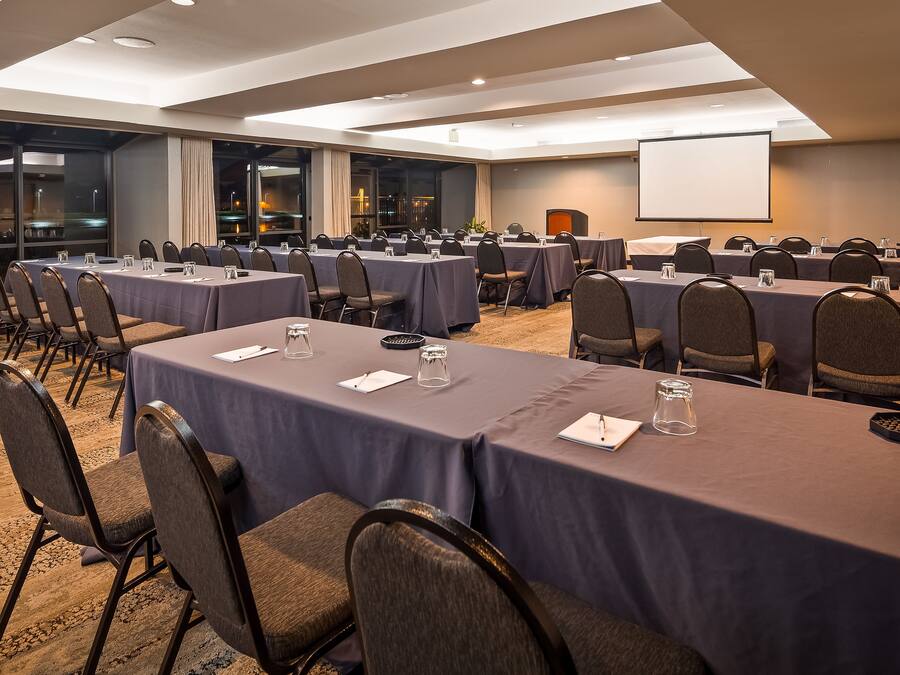 Sala de conferências com várias mesas, cadeiras, tela de projetor e amplas janelas.