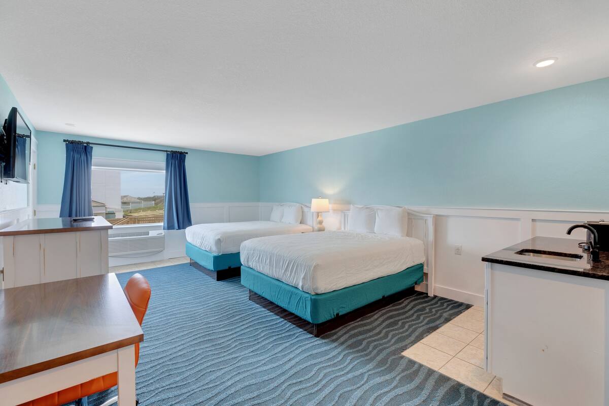 Quarto de hotel com duas camas, uma escrivaninha, uma pia e vista para o exterior.