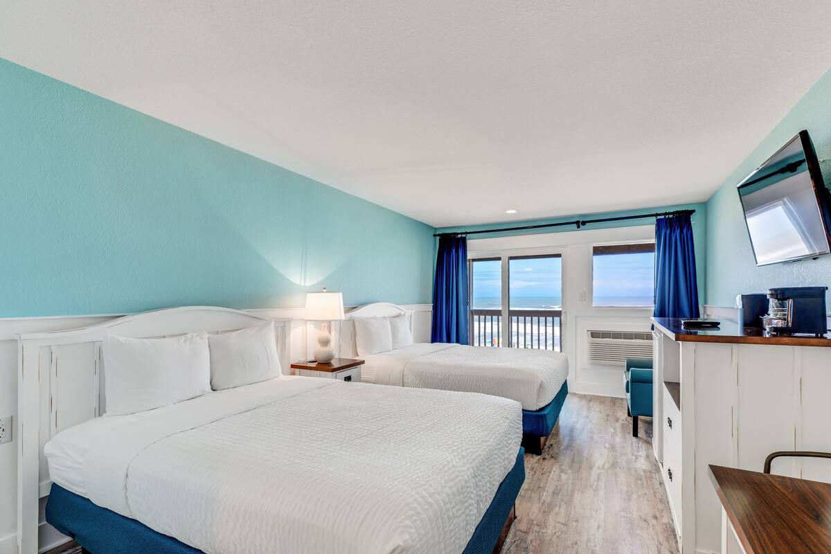 Quarto de hotel com duas camas, uma escrivaninha, uma cadeira e janela com vista para o mar.