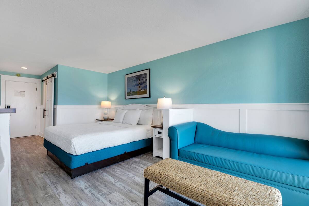 Quarto com cama, sofá azul, mesa de cabeceira com abajures e um quadro na parede.